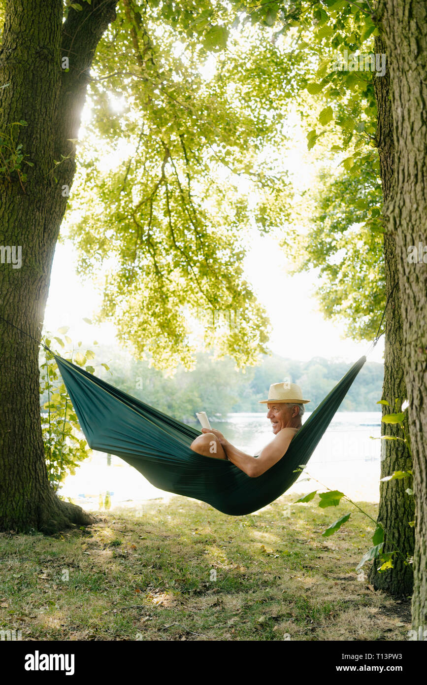 Senior man wearing straw hat relaxing in hammock at lakeshore reading ...