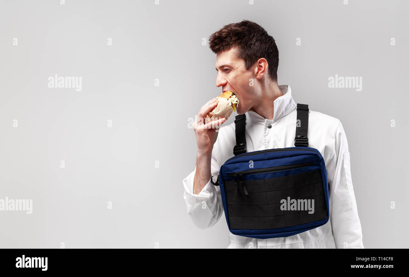 Young stylish male worker with chest rig bag is eating a tasty juicy ...
