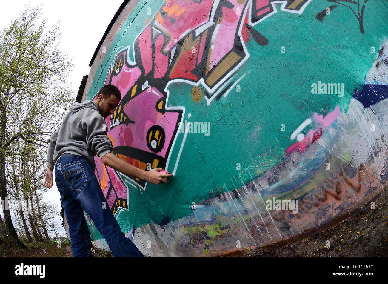 A young guy in a gray hoodie paints graffiti in pink and green colors ...