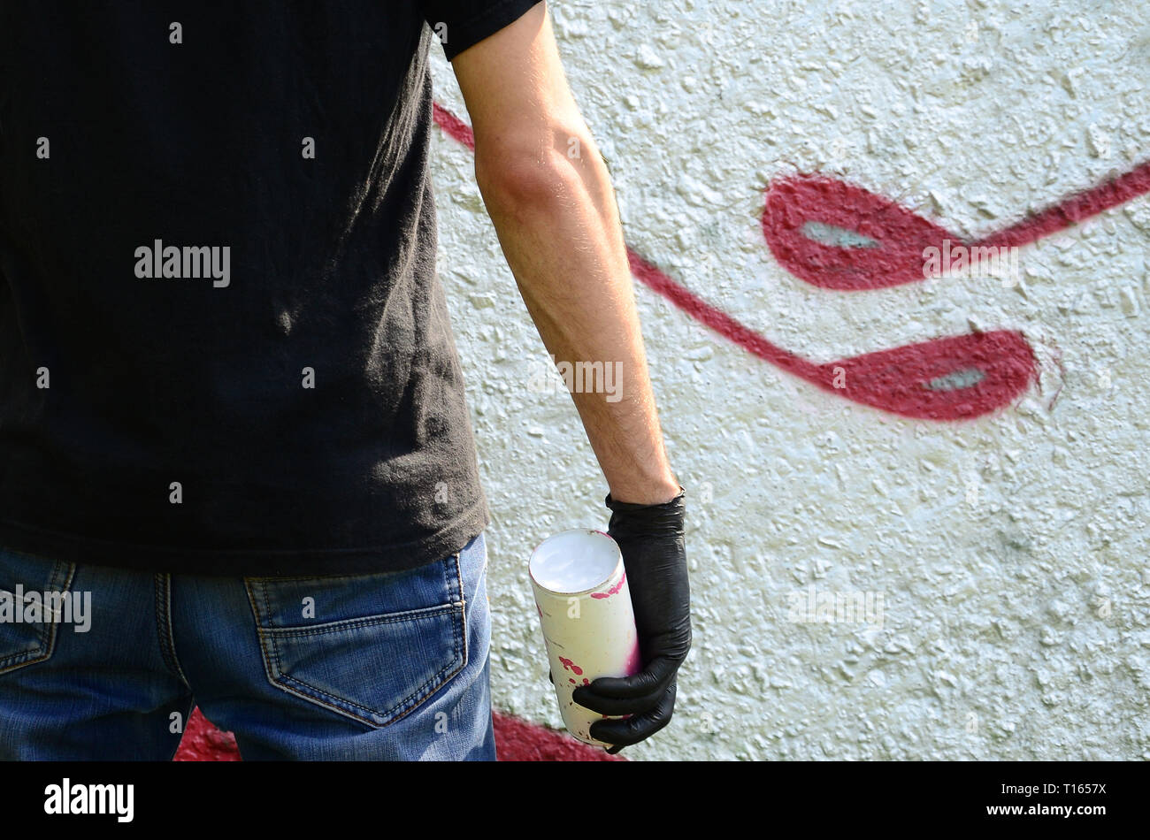 A young hooligan with a spray can stands against a concrete wall with ...
