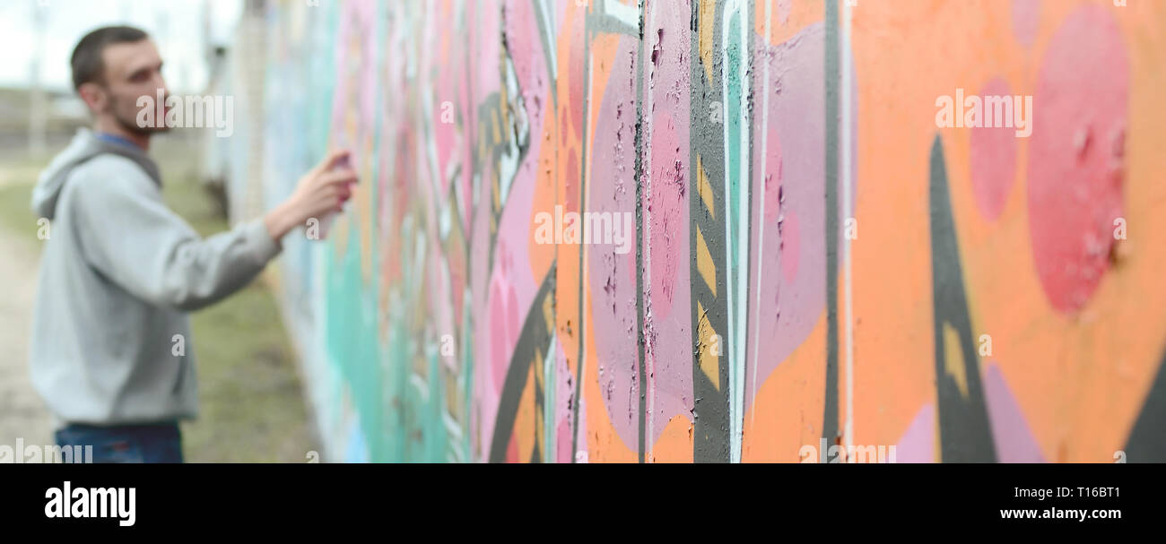 A young guy in a gray hoodie paints graffiti in pink and green colors ...