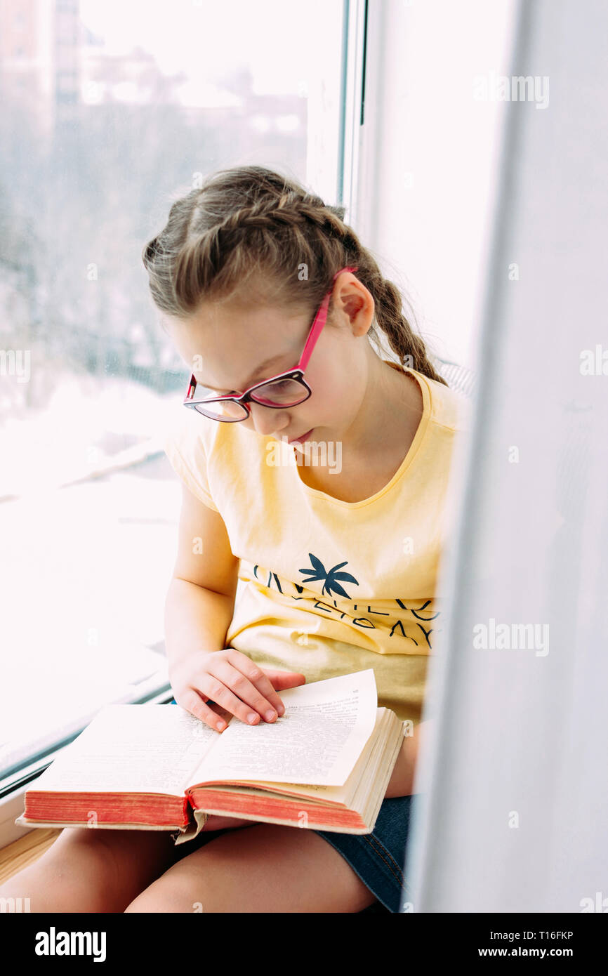 Girl tween with glasses and pigtails reading book at window sill in her ...
