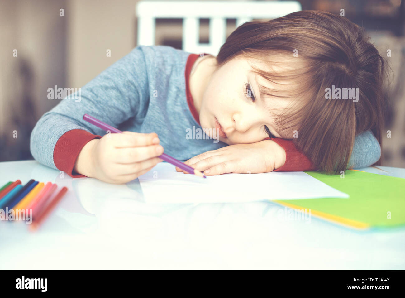A portrait of a tired little girl writing with a pencil on a white ...
