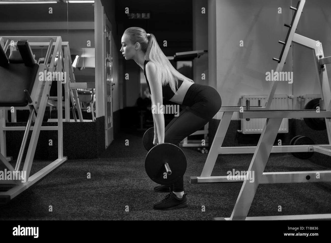 Side view of woman holding heavy barbell and working out in modern, equipped gym. Young, fit and ...