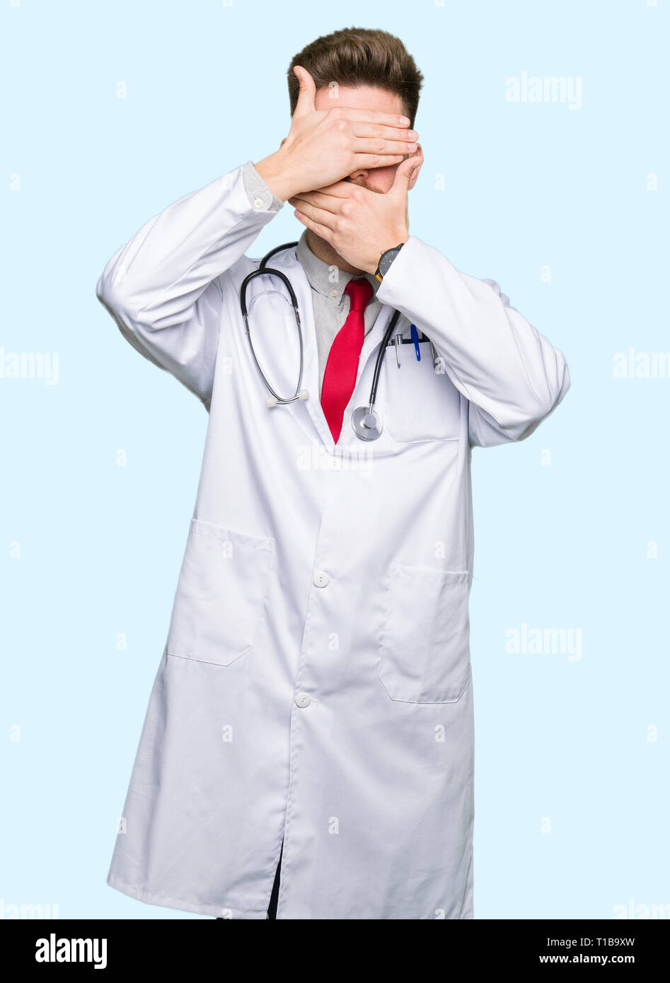 Young handsome doctor man wearing medical coat Covering eyes and mouth ...