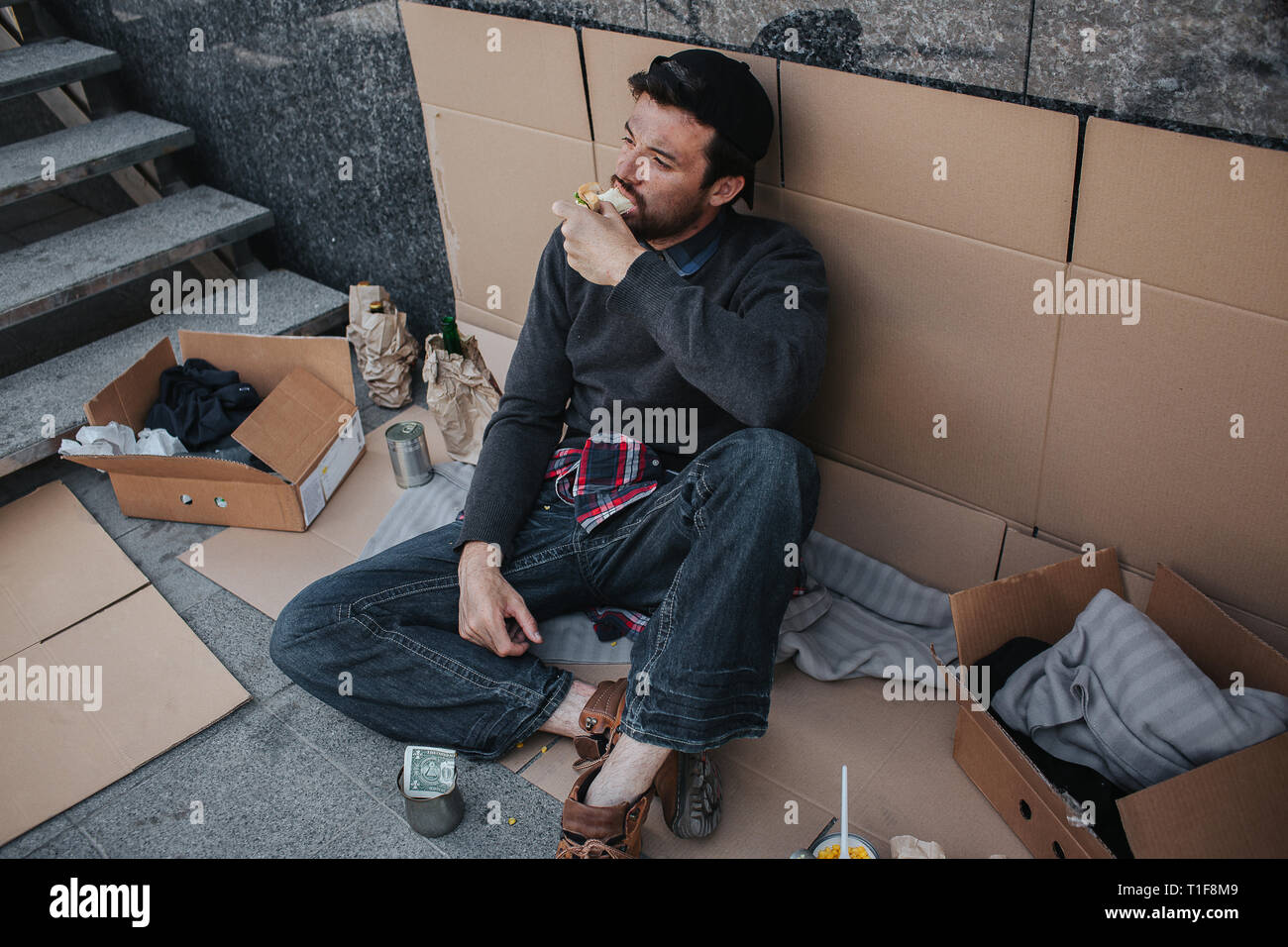 Dark-haired and dirty man is sitting on cardboard and eating sandwich ...