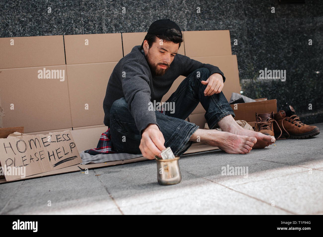 Homeless man is sitting on the cardboard outside and reaching metal cup ...