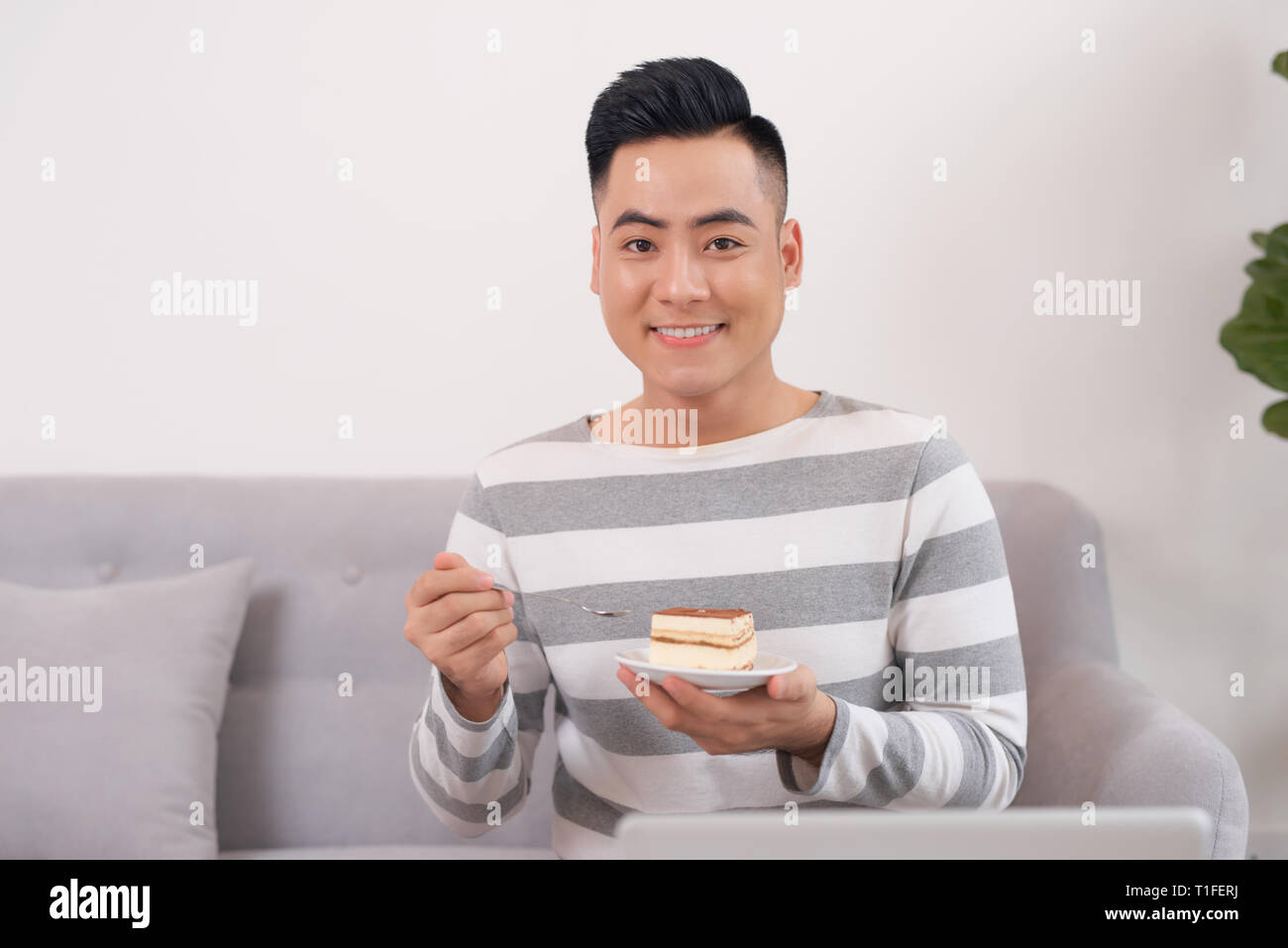 Young asian man eating cake while sitting on sofa at home Stock Photo ...
