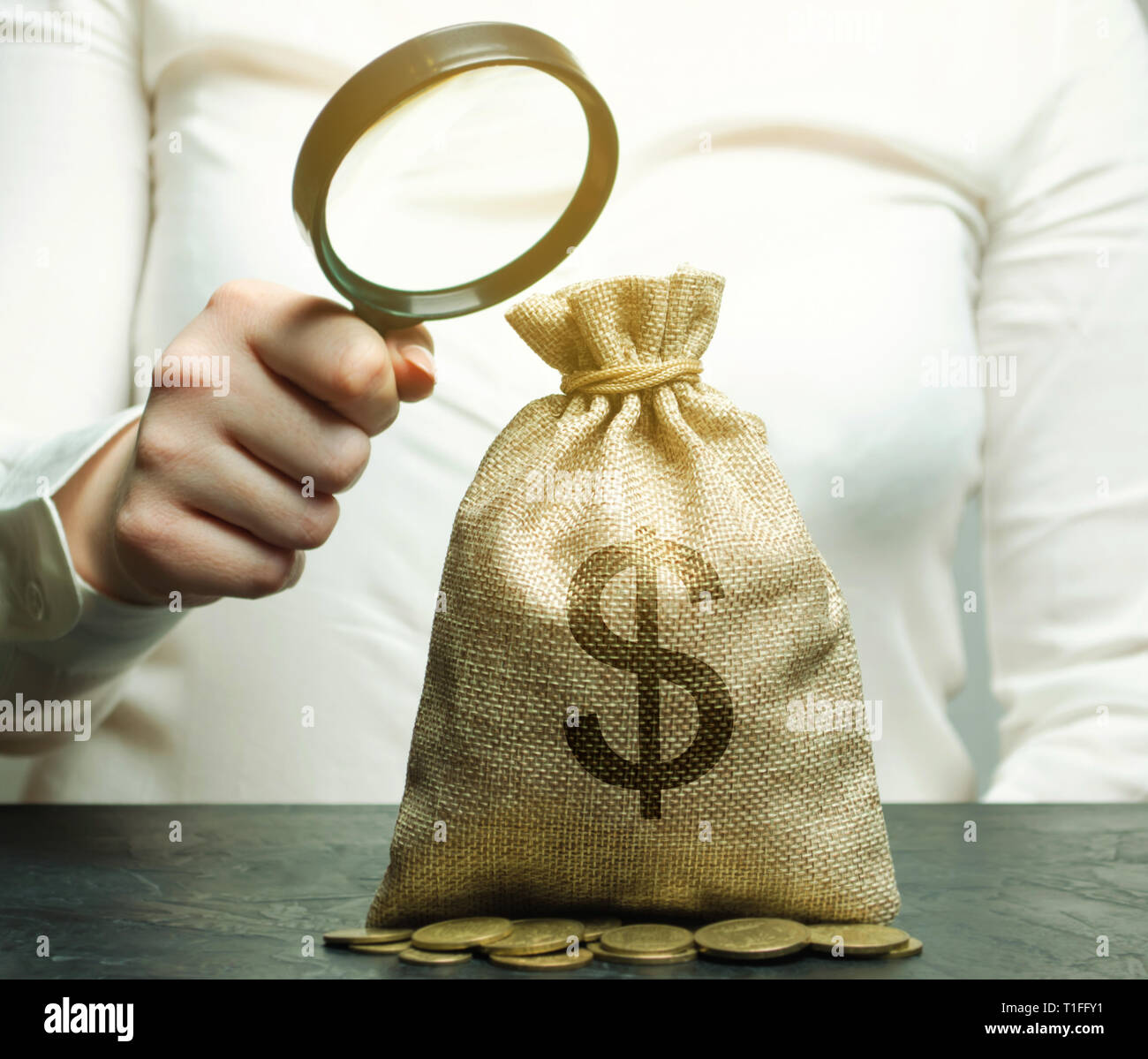 A female hand is holding a magnifying glass over a money bag with coins ...