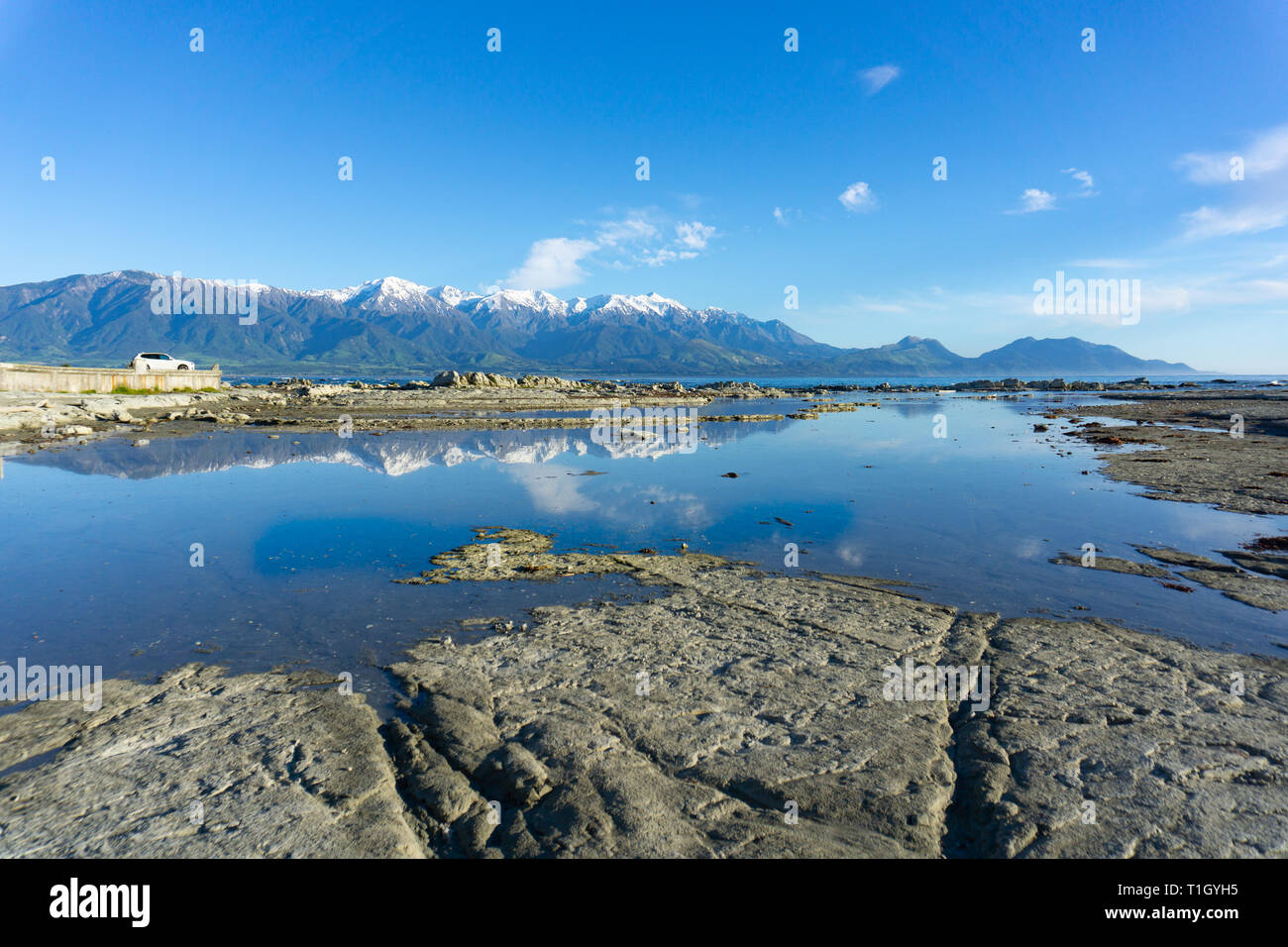 Snow-capped Kaikoura Mountains reflected in calm rockpool on mud-stone ...