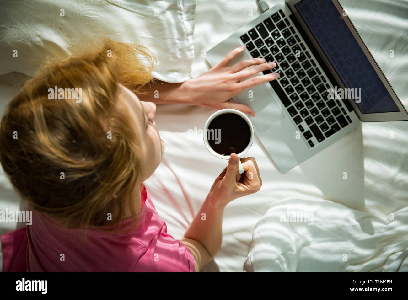 Attractive young woman drinking coffee, lying in bed with laptop ...