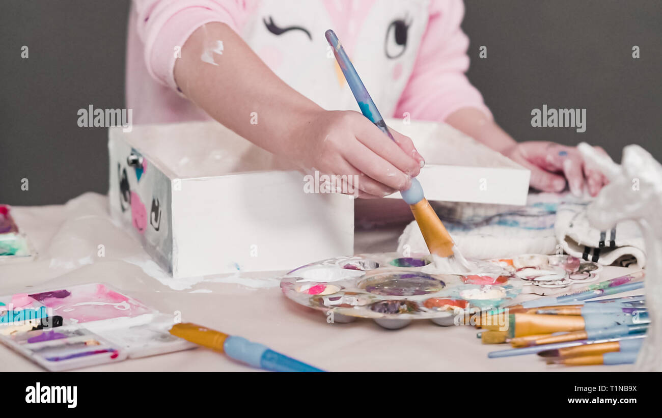 Little girl painting a white unicorn with acrylic paint on a wooden box ...