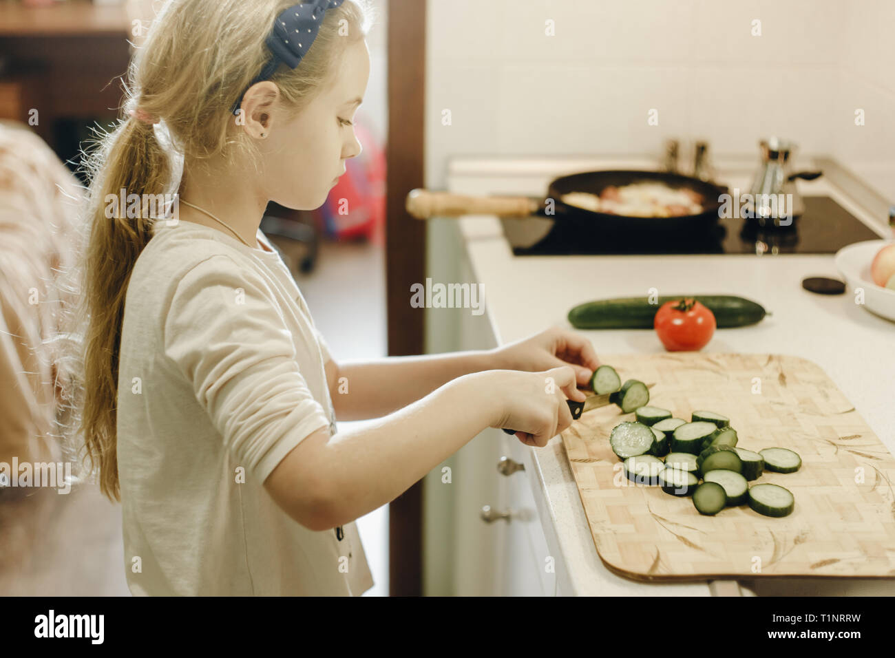 Little blonde girl cutting vegetables while cooking in kitchen at home ...