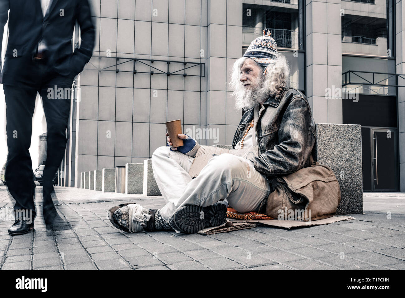 Miserable grey-haired old homeless motionlessly sitting on cold ground ...