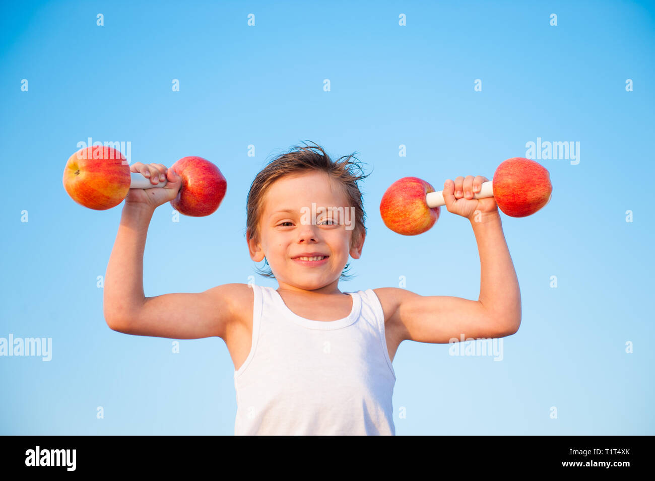 happy healthy little boy lifting apple dumbbells on blue sky background ...