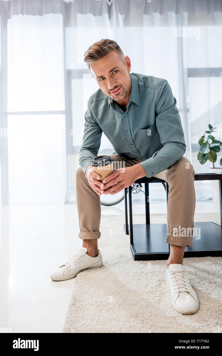 handsome man sitting on coffee table and holding paper cup with drink ...