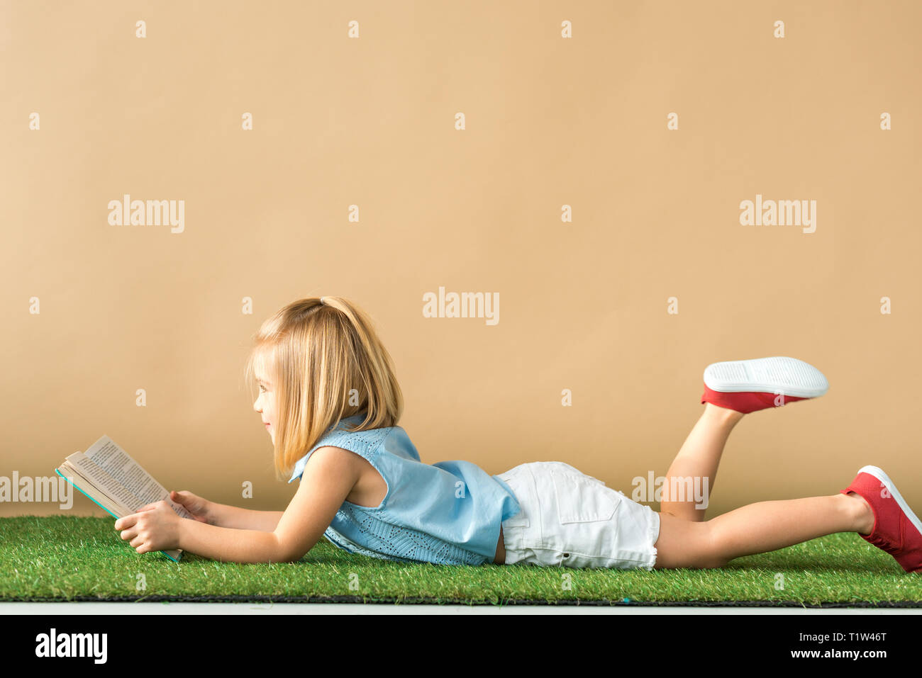 side view of kid lying on grass rug and reading book on beige ...