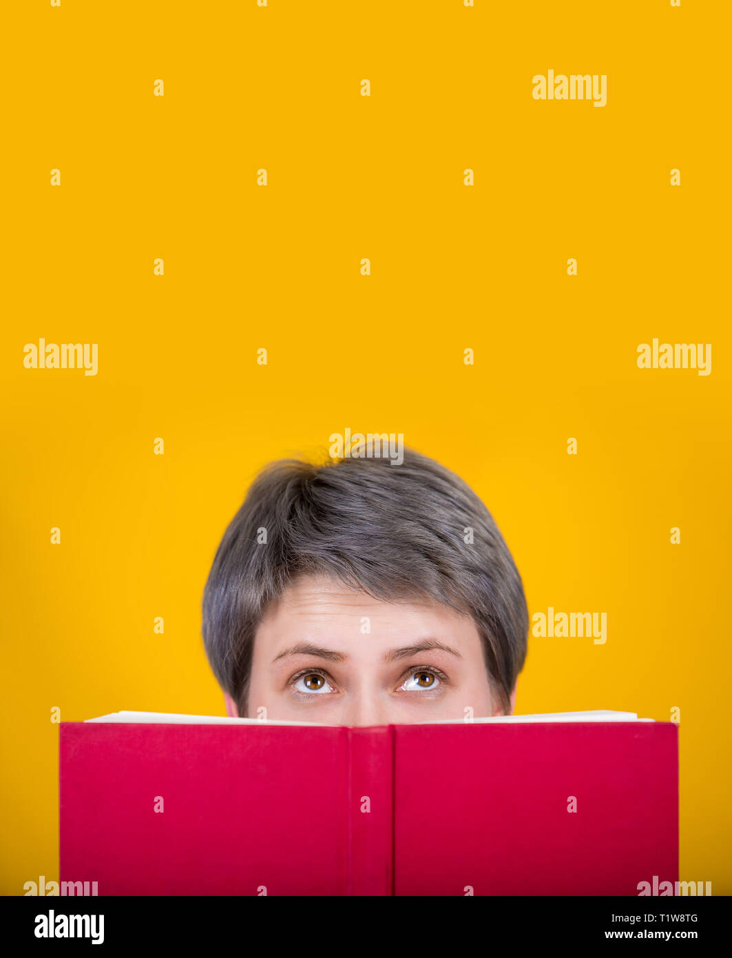 Young woman book reader hides behind a red textbook, looking up has ...