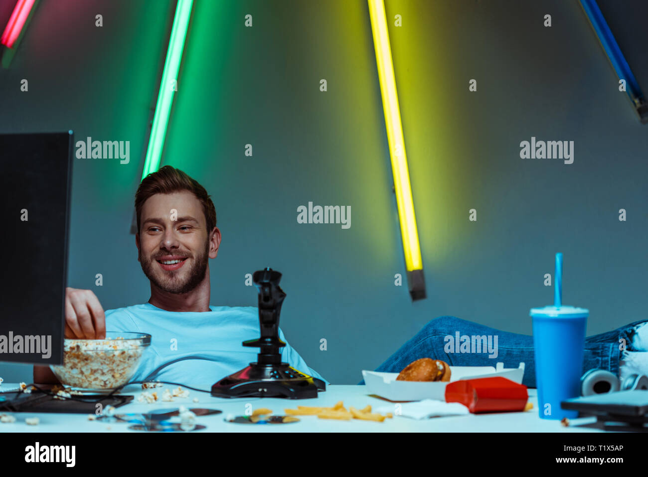 handsome and smiling man looking at computer monitor and eating popcorn ...