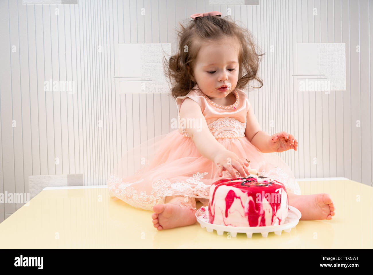 Little baby girl in pink dress sitting on table surface and eating cake ...