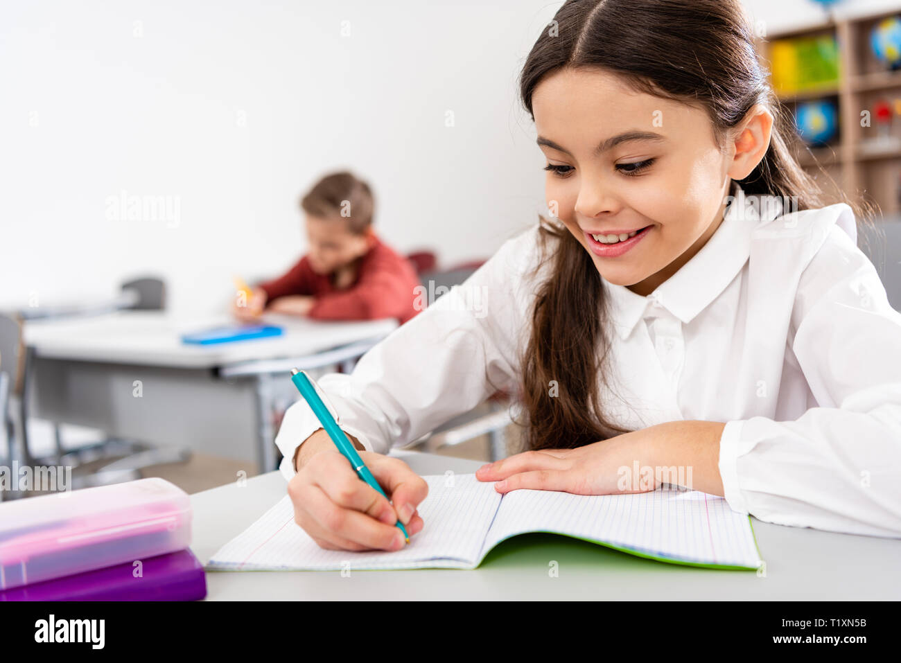 Smiling schoolgirl writing in notebook during lesson in classroom Stock ...
