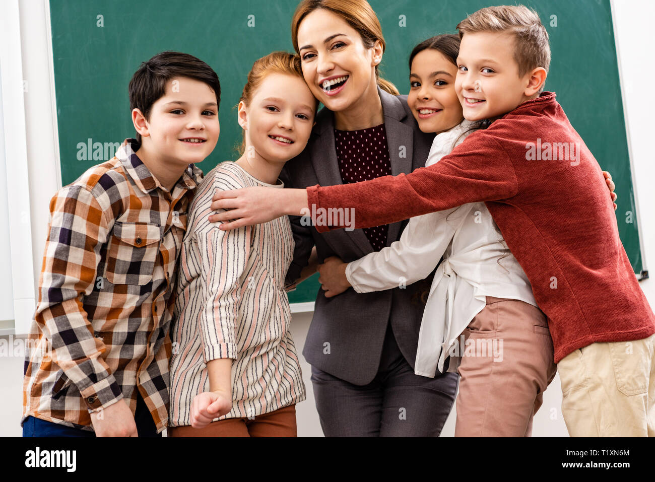 Happy pupils embracing teacher in front of blackboard in classroom ...