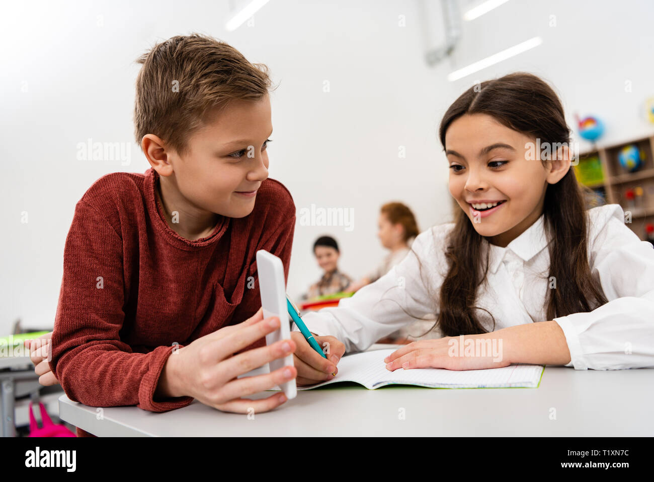Smiling schoolboy showing smartphone to friend at desk in classroom ...