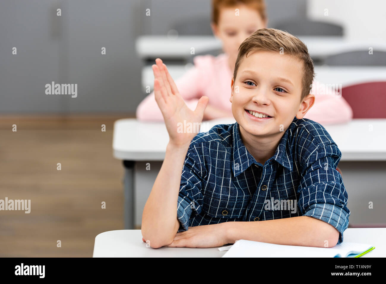 Schoolboy in checkered shirt raising hand up during lesson in classroom ...