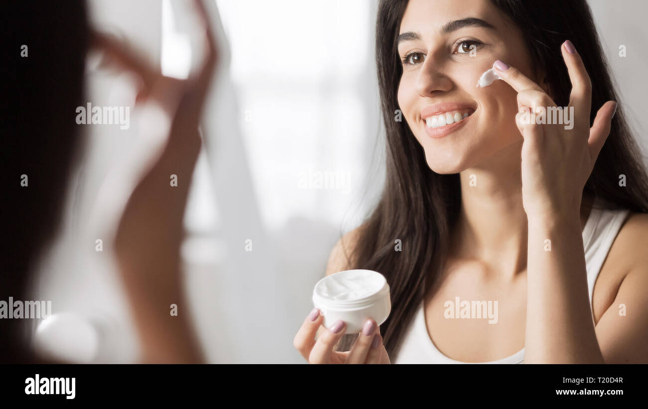 Happy girl applying cream on her face, standing in bathroom, reflecting ...