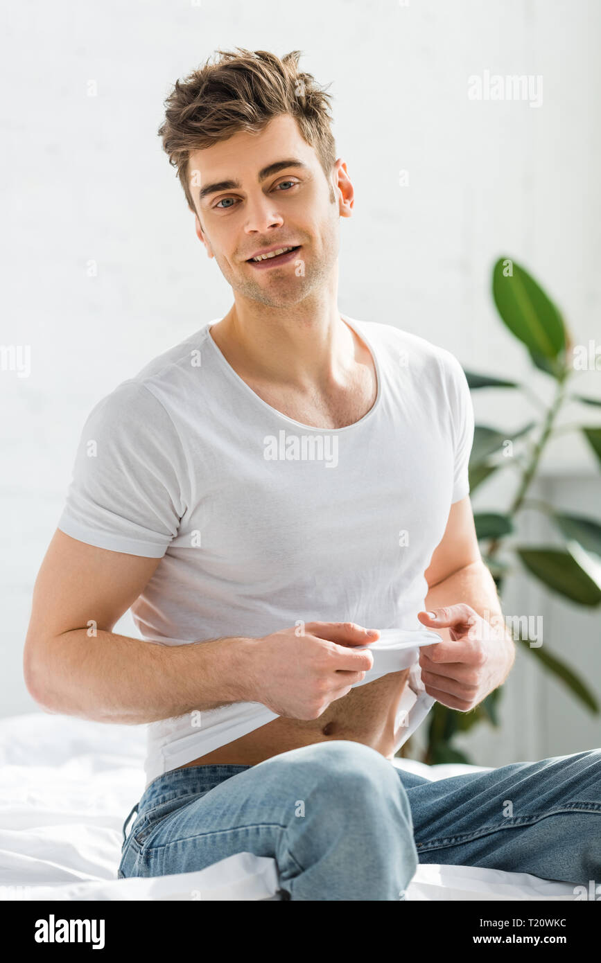 man in jeans sitting on bed and taking off t-shirt in bedroom Stock ...