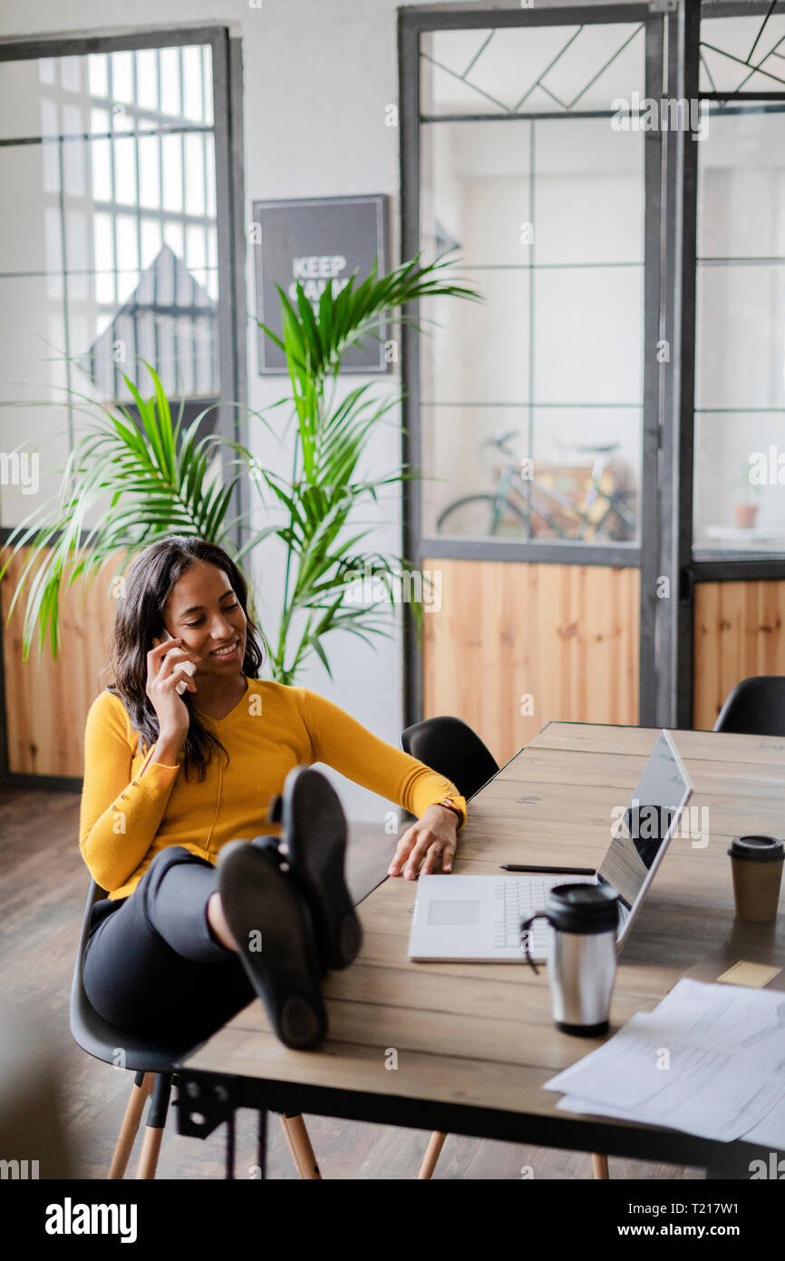 Young businesswoman sitting with feet on desk talking on cell phone ...