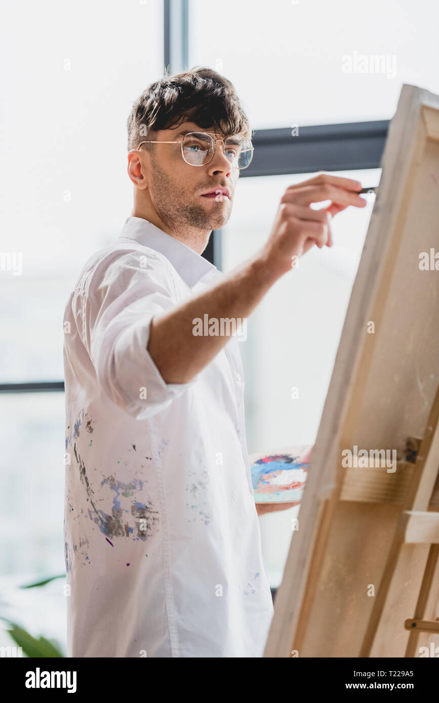 selective focus of handsome artist in glasses painting on canvas Stock ...