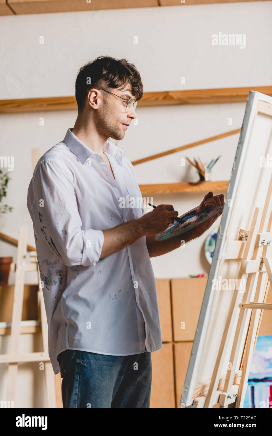 good-looking artist mixing paints on palette while standing near easel ...