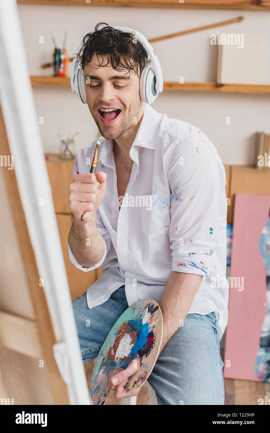 selective focus cheerful artist in headphones, singing with paintbrush ...