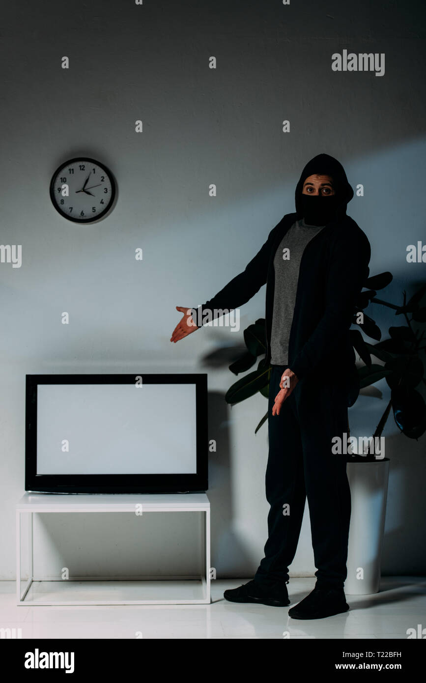 Surprised thief in black mask pointing with hands at flat-screen tv ...