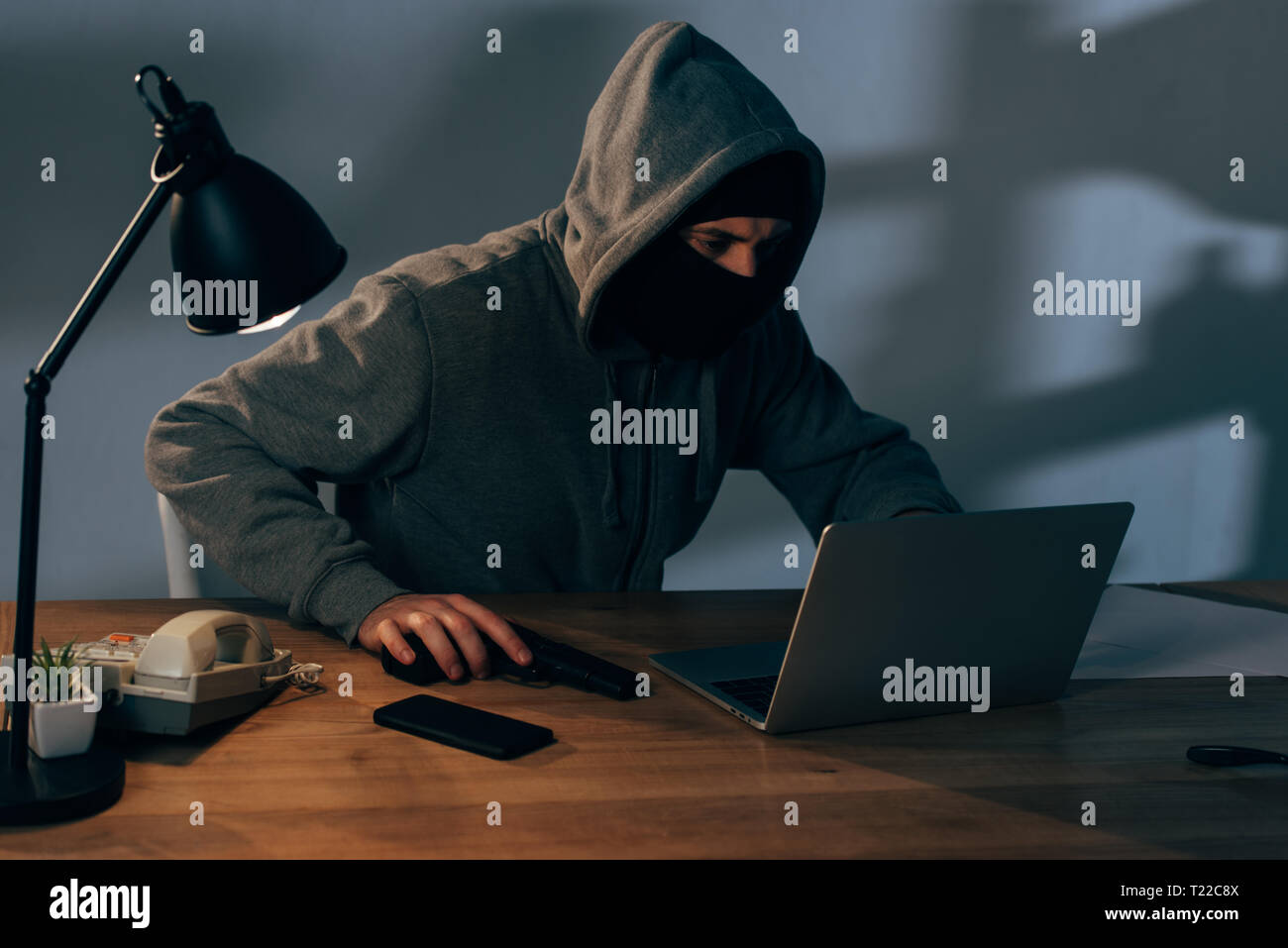 Criminal in mask with gun using laptop in dark room Stock Photo - Alamy