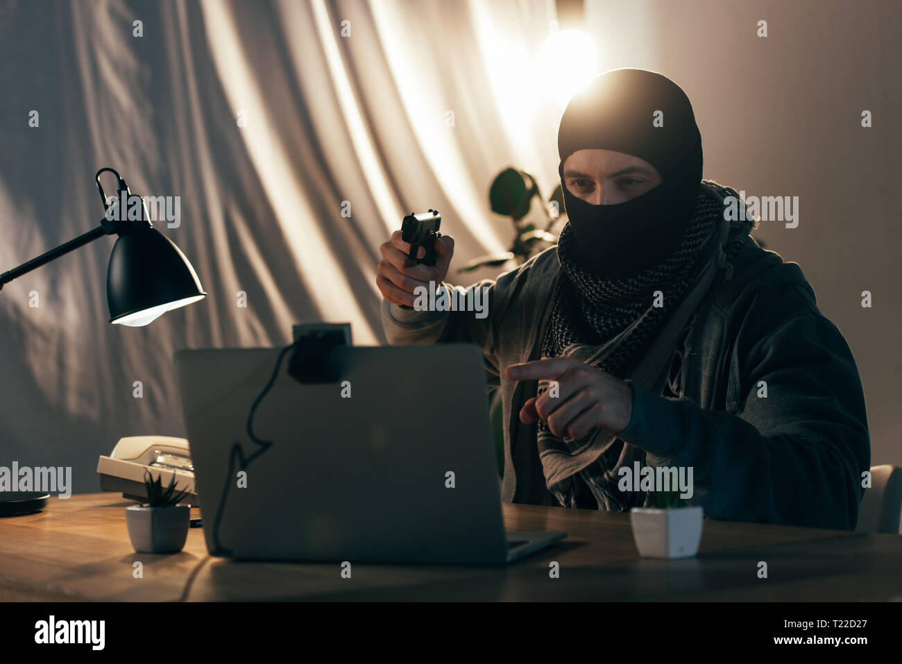 Criminal in mask pointing with finger and aiming gun at laptop screen ...