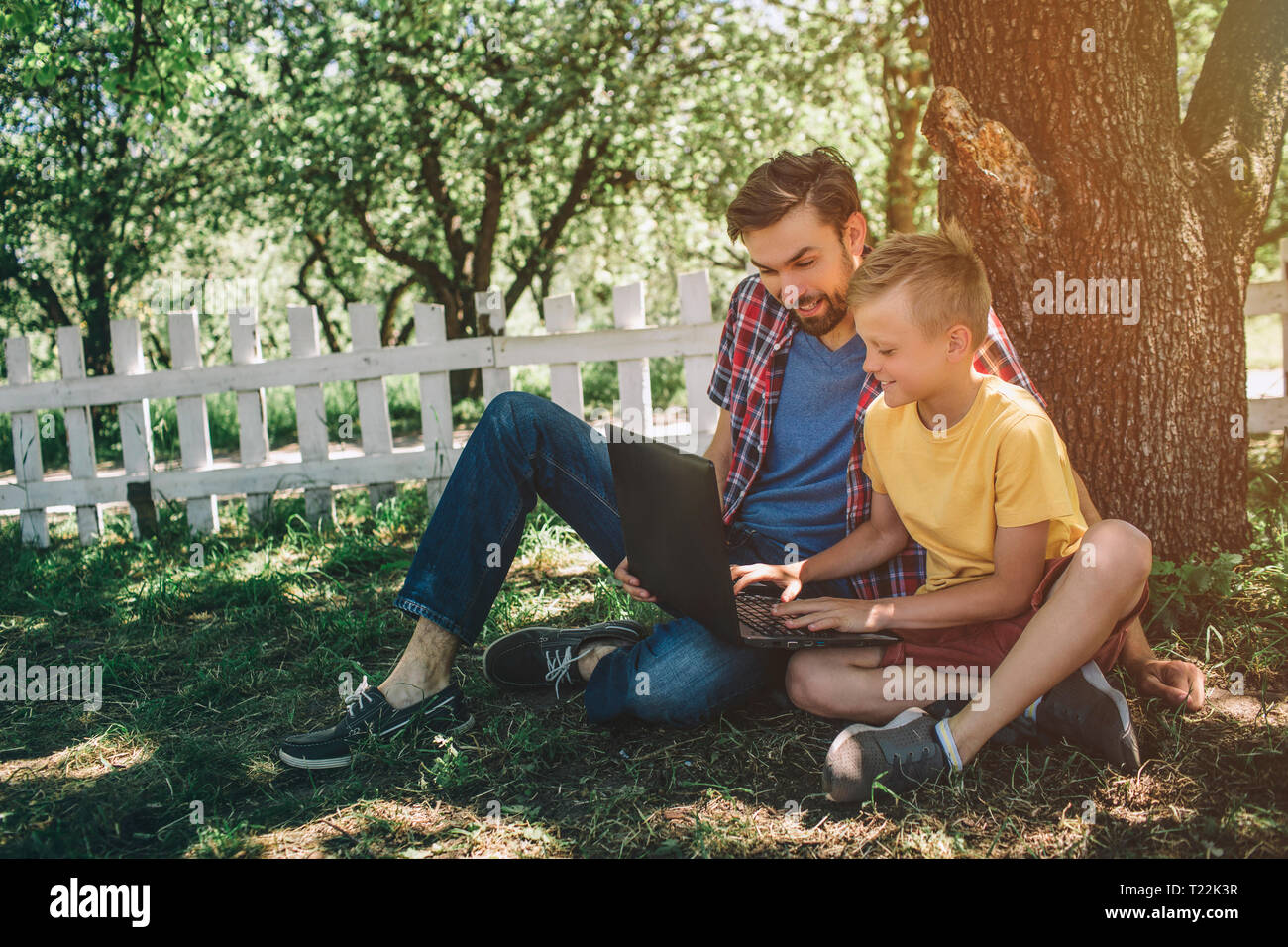 Two people are sitting together under tree and looking at laptop's ...