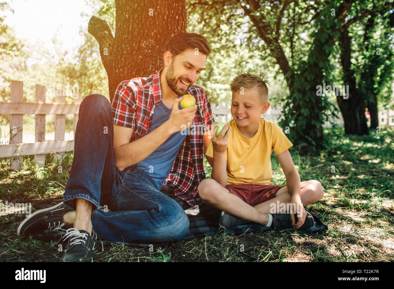 man is sitting on blanket with his son. Guy is looking at child's apple ...