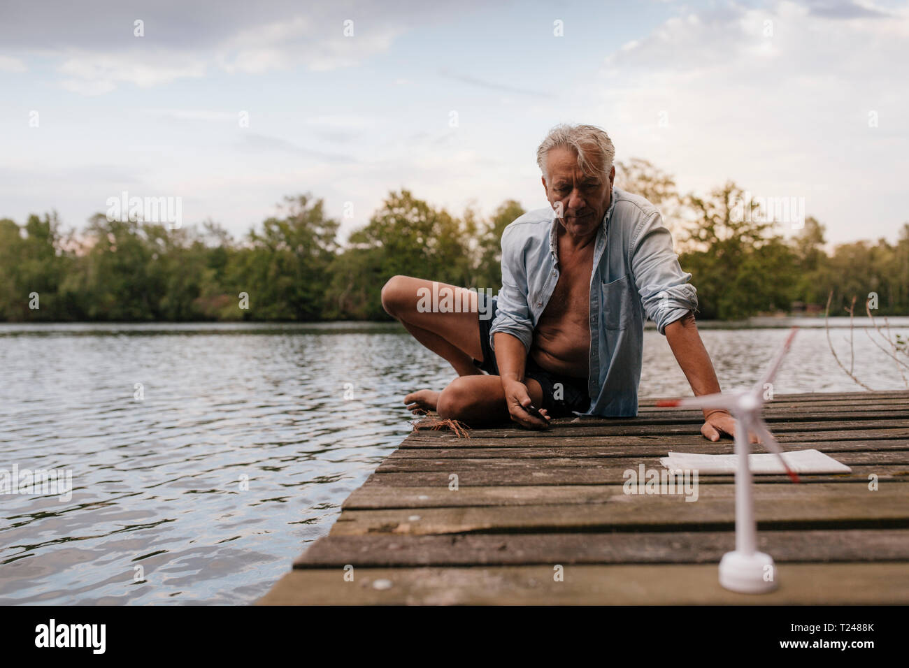 Senior man sitting on jetty at a lake with small wind turbine model ...