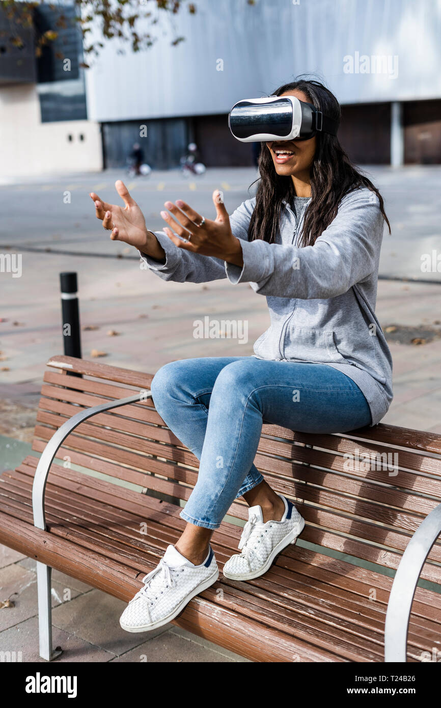 Young woman sitting on a bench, using VR goggles, reaching out with her ...