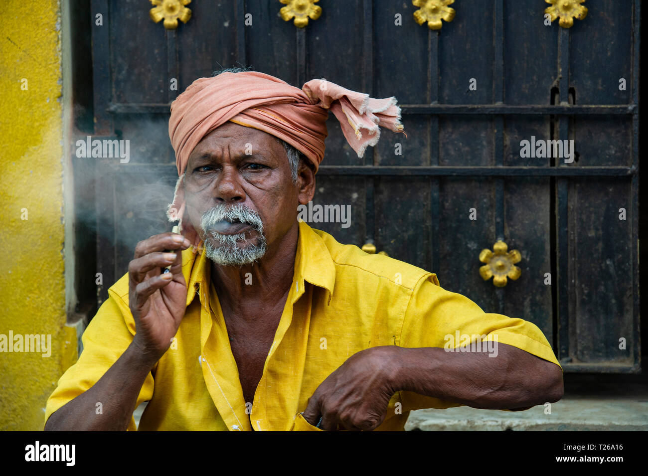 A wise looking man with a turban style hat smokes a cigarette and ...
