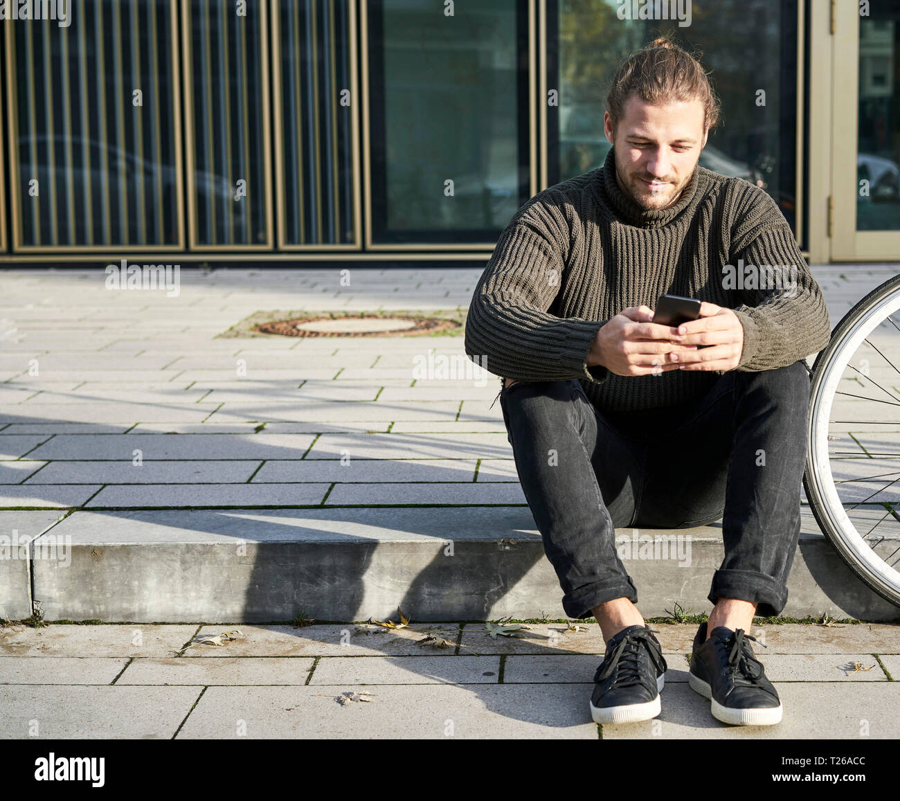 Portrait of bearded young man sitting on curb next to bicycle using ...