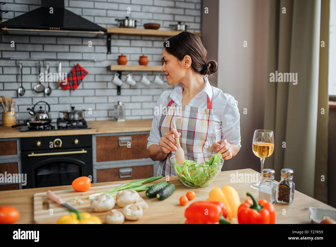 Careful brunette woman sit on table and cook in kitchen. She mix salad ...
