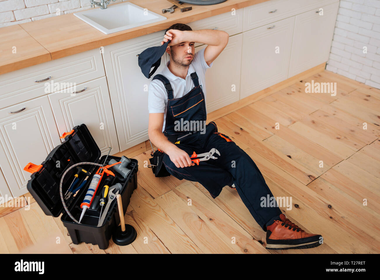 Tired exhausted plumber sit on floor in kitchen. He hold hand on ...