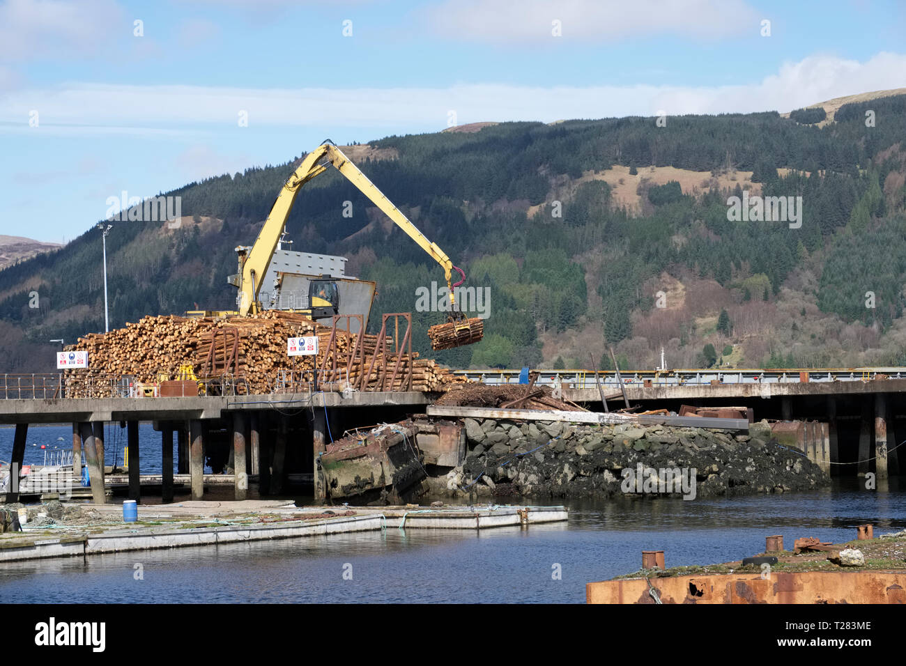 Crane lifting wood logs from stack to ship in sea loch for ...