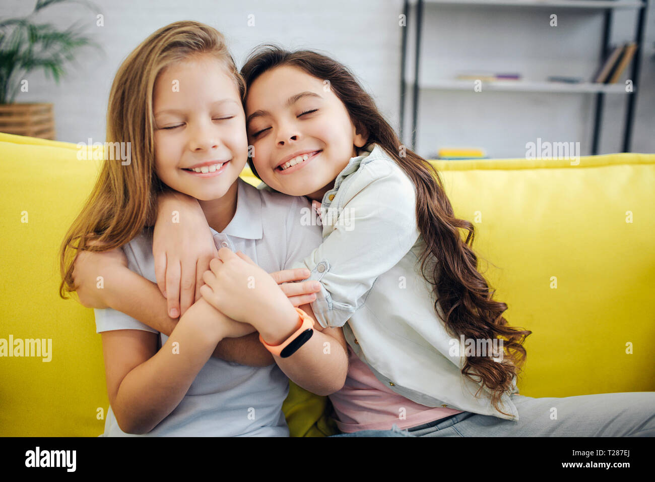 Two cheerful girls sitting on yellow sofa and hug each other. They keep ...