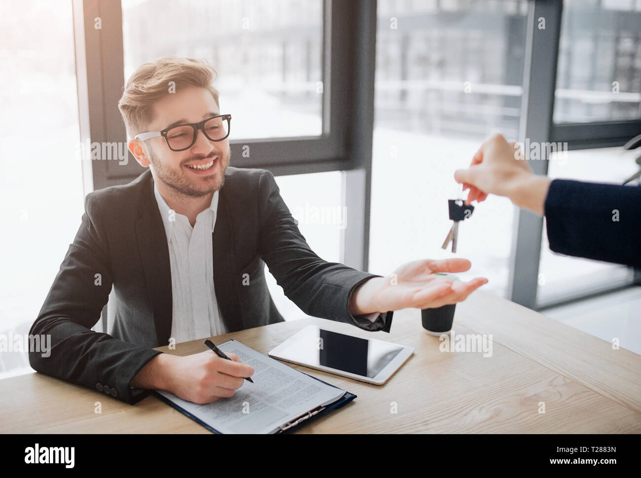 Happy young man in suit sit at table in room. He get keys from another ...