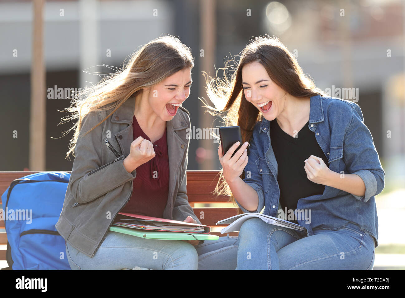 Excited students checking grades on smart phone sitting on a bench in a ...