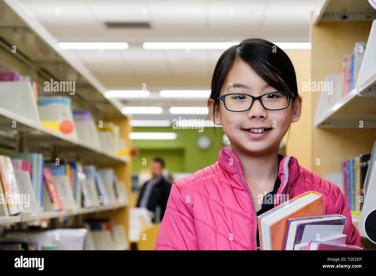 Teenage Asian girl with eyeglasses standing with books in the library ...